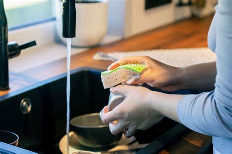 한 여성이 부엌에서 설거지를 하고 있습니다 손에 선택적으로 초점을 맞춥니다 컵과 손에 스폰지를 씻으십시오 창문과 거리 전망이있는 주방 Water Works Park