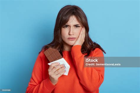 손에 초콜릿의 바를 들고 화가 백인 여성은 과자를 물고 손바닥으로 뺨을 덮고 캐주얼 스웨터를 입고 파란색 배경에 고립 된 후 치통을 느낍니다 의료 20 24세에 대한 스톡