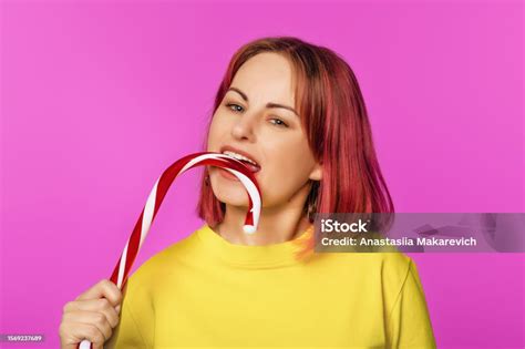 행복한 예쁜 젊은 여자가 달콤한 사탕을 핥고 롤리팝 룩 카메라를 먹는다 분홍색 머리와 분홍색 배경 위에 서 있는 노란색 티셔츠를 입은 사랑스러운 여성 모델 30 34세에