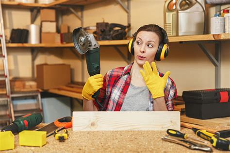 격자 무늬 셔츠에 젊은 갈색 머리 여자 회색 T셔츠 소음 절연 헤드폰 노란색 장갑 다른 도구 나무 테이블 장소에 목공 워크숍에서
