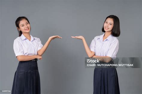 짧은 머리와 긴 머리를 가진 두 명의 십대 아시아 여학생 교복을 입고 중앙에 빈 공간을 제시하는 손을 들고 밝게 웃으며 회색 배경 스튜디오에서 사진을 찍었다 16 17 살에