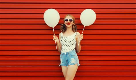 하얀 하트 모양의 선글라스를 끼고 빨간 배경에 반바지를 입고 풍선을 들고 즐거운 시간을 보내는 행복한 쾌활한 젊은 여성 25 29세에 대한 스톡 사진 및 기타 이미지