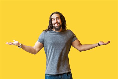 몰라요 회색 티셔츠에 긴 곱슬 머리를 하고 머리를 길게 하고 두 팔을 들고 혼란스러운 얼굴로 카메라를 바라보는 수염을 기른 청년의 모습 질문하기에 대한 스톡 사진 및 기타