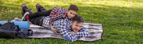 Happy Young Father Resting In Park And Small Son Lying On Back Of Dad
