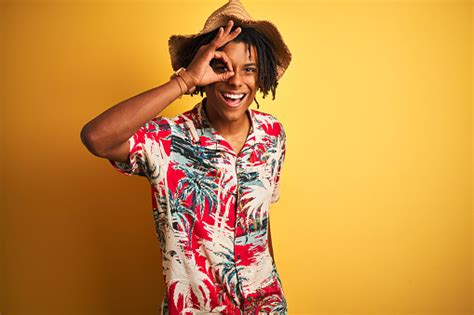 꽃 무늬 셔츠와 모자를 쓰고 고립 된 노란색 배경 위에 꽃을 입은 아프로 미국 남자는 행복한 얼굴로 손가락을 통해 보는 손으로 미소짓는 확인 제스처를하고 있습니다 구멍에