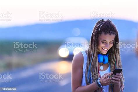 일몰에 자동차와 해변 도로에 그녀의 휴대 전화와 함께 서 꼰 긴 머리와 미소 젊은 여자 휴대전화에 대한 스톡 사진 및 기타 이미지 휴대전화 십대 휴대용 통신 기기