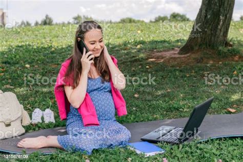 긍정적 인 아름다운 검은 머리 임신 한 여성은 노트북과 잔디밭에 공원에 앉아 전화로 이야기 모성 및 임신 개념 임산부를 위한 원격 작업의 개념 30 34세에 대한 스톡 사진