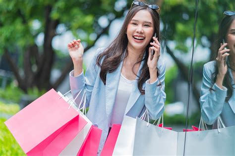 매력적인 아시아 여자는 긍정적 인 캐주얼 쇼핑 핸드 홀드 종이 봉지와 스마트 폰을 기쁘게 구매 천을 즐길 수 있습니다 구매 천 쇼핑 센터 몰 상점 앞 복도 백화점에서 판매