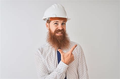 고립된 흰색 배경 위에 보안 헬멧을 쓰고 있는 젊은 빨간 머리 아일랜드 건축가 남자는 얼굴에 행복하고 자연스러운 표정으로 손과 손가락으로 가리키는 얼굴의 미소와 함께