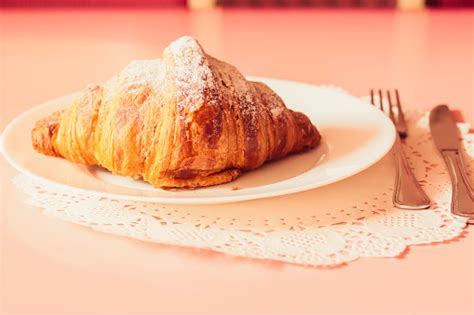 흰색 접시에 아이싱 슈가가 뿌려진 프렌치 아몬드 크루아상 분홍색 테이블에 포크와 나이프가 담긴 아침 식사 제공 건강한 생활방식에