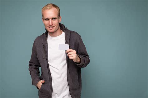 회색 스웨터와 흰색 티셔츠를 입은 잘 생긴 자신감금발 남자의 사진은 파란색 배경 벽에 고립 되어 있고 카메라를 바라보는 신용 카드를 들고 있다 T 셔츠에 대한 스톡 사진 및