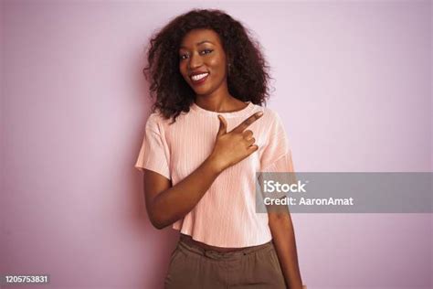 고립된 분홍색 배경 위에 서있는 티셔츠를 입은 젊은 아프리카 계 미국인 여성은 얼굴에 행복하고 자연스러운 표정으로 손과 손가락으로 가리키는 얼굴의 미소와 함께 명랑합니다 T