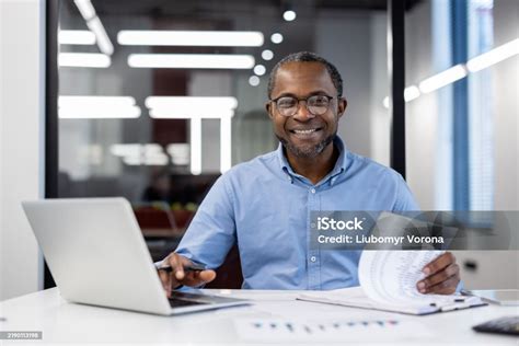 성숙한 아프리카 계 미국인 사업가는 현대적인 사무실 환경에서 노트북으로 작업하면서 열정적으로 중요한 문서를 들고 자신있게 미소 짓고 있습니다 개성 개념에 대한 스톡 사진 및