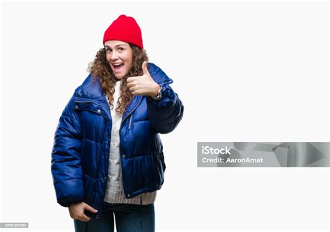 아름 다운 젊은 갈색 머리 곱슬 머리 소녀 행복 엄지손가락 손으로 제스처를 하 고 격리 된 배경 위에 겨울 코트 모직 모자와 스웨터를 입고 승인 성공을 보여주는 카메라를