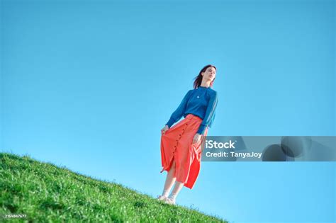 푸른 하늘을 배경으로 풀이 무성한 언덕에 서 있는 파란 상의와 산호 치마를 입은 십대 소녀 밝은 색상과 맑은 하늘을 가진 야외 인물 사진입니다 14 15 살에 대한 스톡