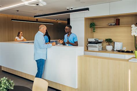 고령의 남성 환자가 친절한 남성 간호사의 도움으로 접수 데스크에 서서 의료 양식을 작성합니다 25 29세에 대한 스톡 사진 및