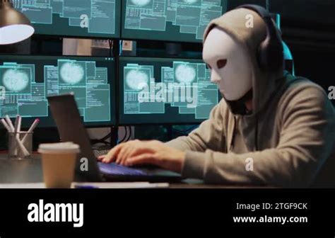 Masked Cyber Thief Hacking Computer Network At Night Working On Laptop And Multiple Monitors To