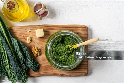 케일 잎 호두 파마산 치즈 마늘 올리브 오일과 같은 기본 재료가 들어간 흰색 나무 표면 위에 도마에 숟가락이 달린 유리 그릇에 담긴 수제 케일 페스토 0명에 대한 스톡 사진