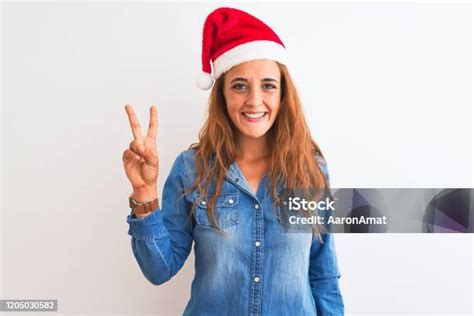 고립 된 배경을 통해 크리스마스 모자를 쓰고 있는 젊은 아름다운 빨간 머리 여성은 자신감과 행복한 미소를 지으면서 손가락 번호 2를 가리키고 있습니다 가리키기에 대한 스톡