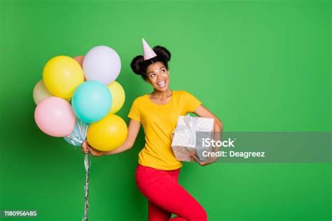 많은 공기 풍선과 큰 기프트 박스를 들고 예쁜 어두운 피부 아가씨의 사진은 캐주얼 노란색 티셔츠 빨간 바지 종이 모자 고립 된 녹색 배경을 입고 빈 공간을 찾고 갈색 머리에