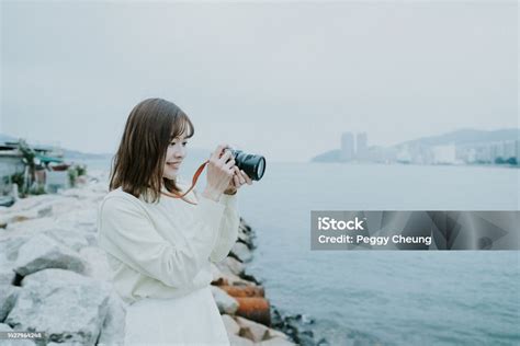 젊은 아시아 여성 사진 작가가 바위가 많은 해변과 해안선에서 바다와 물 건너편에있는 도시 앞에서 사진을 찍습니다 20 24세에 대한 스톡 사진 및 기타 이미지 Istock