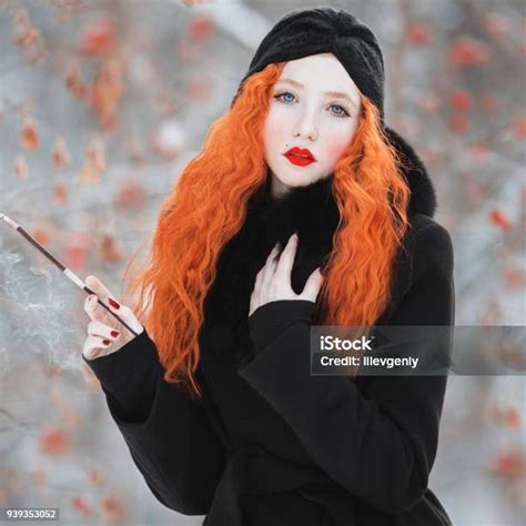 손에서 떠 있는 겨울 숲의 배경에 검은 코트에 붉은 머리를 가진 여자 담배와 그녀의 머리에 터 번으로 밝은 모습으로 빨간 머리 소녀 흡연 미학 감각 지각에 대한 스톡 사진