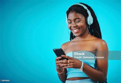 파란색 배경에 서있는 동안 헤드폰을 착용하고 휴대 전화를 사용하는 젊은 여성의 샷 사람들에 대한 스톡 사진 및 기타 이미지