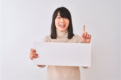 젊은 아름다운 중국 여자는 행복한 얼굴 번호 하나 손가락을 가리키는 아이디어 또는 질문으로 놀란 고립 된 흰색 배경 위에 서있는 배너를 들고 가리키기에 대한 스톡 사진 및