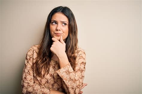 젊은 아름 다운 갈색 머리 여자 캐주얼 티셔츠를 입고 흰색 배경 위에 서 턱에 손으로 질문에 대 한 생각 펜시형 표정 사려 깊은 얼굴로 미소 의심 개념 T 셔츠에 대한 스톡