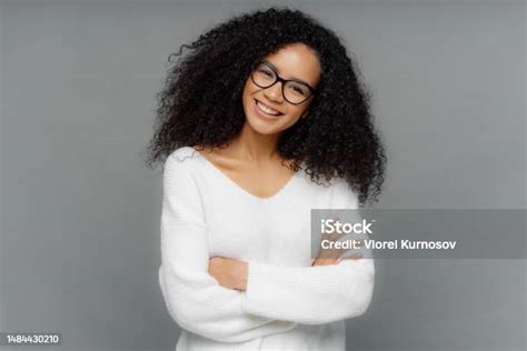 친절 한 찾고 아프리카계 미국인 여자의 절반 길이 촬영 머리를 틸트 어두운 상쾌한 덥 머리 가슴 위에 접힌 팔을 유지 캐주얼 흰색 스웨터를 착용 스튜디오에서 사진 촬영을