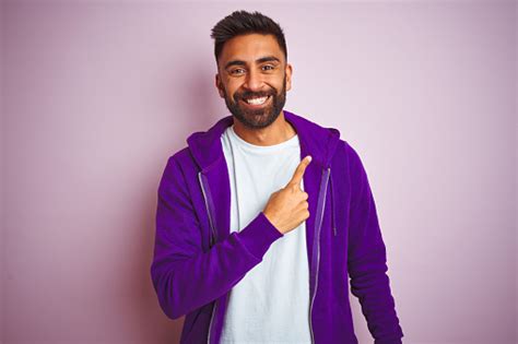 보라색 스웨트셔츠를 입은 젊은 인도 남자가 고립 된 분홍색 배경 에 서서 행복하고 자연스러운 표정으로 손과 손가락으로 옆으로 가리키는 얼굴의 미소로 명랑합니다 가리키기에