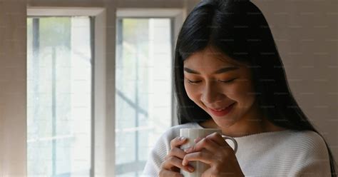 하얀 면 드레스를 입은 젊은 창조적인 여성이 커피 컵을 손에 들고 편안한 거실을 배경으로 나무 테이블에서 일하는 사진 사진