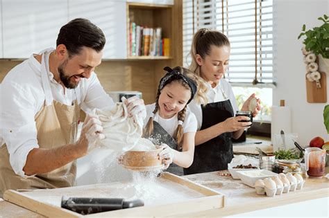 가족이 함께 즐거운 시간을 보내고 있습니다 딸과 행복한 아버지가 나무 표면에 밀가루를 체질합니다 금발 엄마가 잼을 엽니 다 나무 테이블에 준비된 재료 큰 창문이있는 현대적인