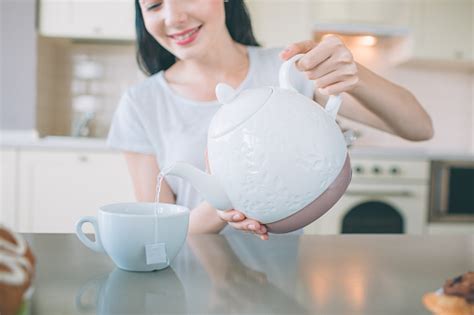 테이블에 앉아 갈색 머리의 절단 보기 그녀는 흰색 주전자를 들고 컵에 물을 붓는 다 차를 준비 하 고 있는 소녀 그녀는 부엌에서 Sitd 가정 생활에 대한 스톡 사진 및