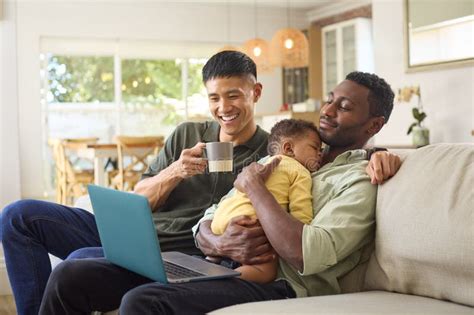 Same Sex Male Couple With Sleeping Baby Babe On Sofa At Home Working On Laptop Stock Image