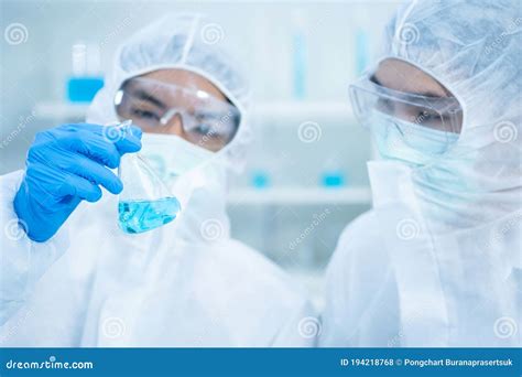 Two Young Scientists In Virus Protection Suit And Wear Hygiene Mask Seeing Blue Solution In The
