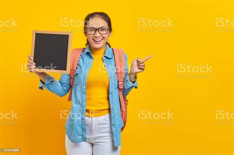 배낭을 들고 캐주얼 한 옷을 입은 젊은 아시아 여성 학생의 초상화는 빈 칠판을 들고 노란색 배경에 고립 된 손가락으로 복사 공간을 가리키고 있습니다 25 29세에 대한 스톡