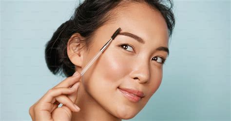 아름다움 웃는 여자는 메이크업 브러쉬 클로즈업으로 눈썹을 닦는다 아름다운 행복한 아시아 소녀 모델의 초상화는 메이크업을 하고 스튜디오에서 화장품 브러시로 눈썹을