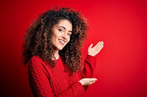 곱슬 머리와 피어싱 캐주얼 빨간 스웨터를 입고 젊은 아름다운 여자는 열린 손으로 자연 미소를 입력 초대 들어가기에 대한 스톡