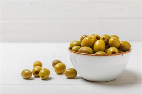 มะกอกสีเขียวบนพื้นหลังไม้สีขาว มะกอกชนิดต่าง ๆ ในชามและน้ํามันมะกอกที่มีใบมะกอกสด พื้นที ภาพ