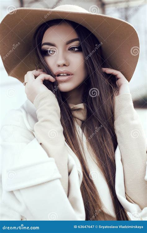 Девушка очарования с темными прямыми волосами носит роскошное бежевое пальто с элегантной шляпой