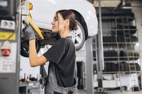 자동차 수리점에서 차 밑을 수리하는 백인 정비사 여성 회색 유니폼을 입은 여성 노동자가 수리 차고에서 자동 리프트로 차를 들어 올립니다 25 29세에 대한 스톡 사진 및