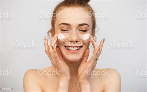 빨간 머리와 주근깨가 있는 아름다운 여인이 하얀 벽에 눈을 감고 웃고 있는 그녀의 얼굴에 두 핸드 크림을 들고 노는 모습을 보며 눈을 감고 있다 주근깨에 대한 스톡 사진 및