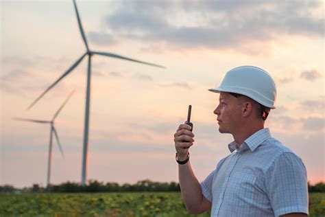 풍력 발전 엔지니어가 풍력 터빈 배경에서 휴대용 무선 송신기와 대화를 나누고 있습니다 헬멧을 쓰고 있는 한 남자가 전기 풍차의 작동을 감독합니다 개발에 대한 스톡 사진 및