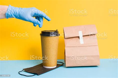 비접촉식 음식 배달 개념 파란색 고무 장갑을 낀 택배 의 손은 주문 보호 얼굴 마스크 커피 한 잔 및 노란색 배경에 종이 봉투를