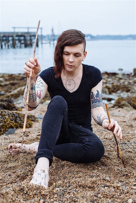 Transgender Gender Fluid Person With Tattoos Sitting On Beach With Stick By Stocksy
