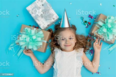 선물과 색종이가 있는 축제 모자를 쓰고 파란 배경에 있는 면흰색 드레스를 입은 달콤한 행복한 어린 소녀가 생일을 축하하는 것을 기뻐하며 텍스트의 장소입니다 6 7 살에 대한