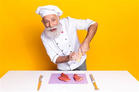 노란 배경의 부엌에서 고기를 요리하는 미소 짓는 수석 요리사 요리에 향신료를 추가 전문 유니폼을 입고 55 59세에 대한 스톡 사진 및 기타 이미지 Istock