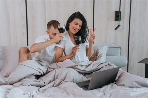 와인 잔을 들고 침대에 앉아 허니문 동안 호텔 방에서 노트북으로 영상 통화를 하고 인터넷을 통해 친구들과 이야기하는 장난기 많은 유럽 커플 행복한 신혼 여행 휴가 기념일 2