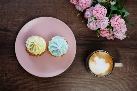 분홍색 접시에 두 개의 노란색과 파란색 크림 캡케이크가 나무 테이블에 앉아 있는 평면도 베이지색 카푸치노 컵이 테이블 위에 놓여